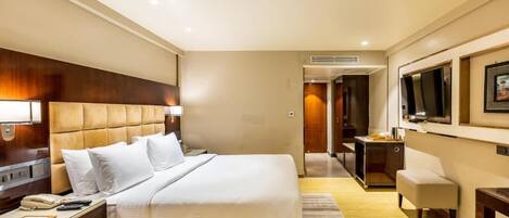 Quarto de hotel com uma cama grande, uma escrivaninha, uma cadeira, televisão e casa de banho visível através de uma porta aberta.