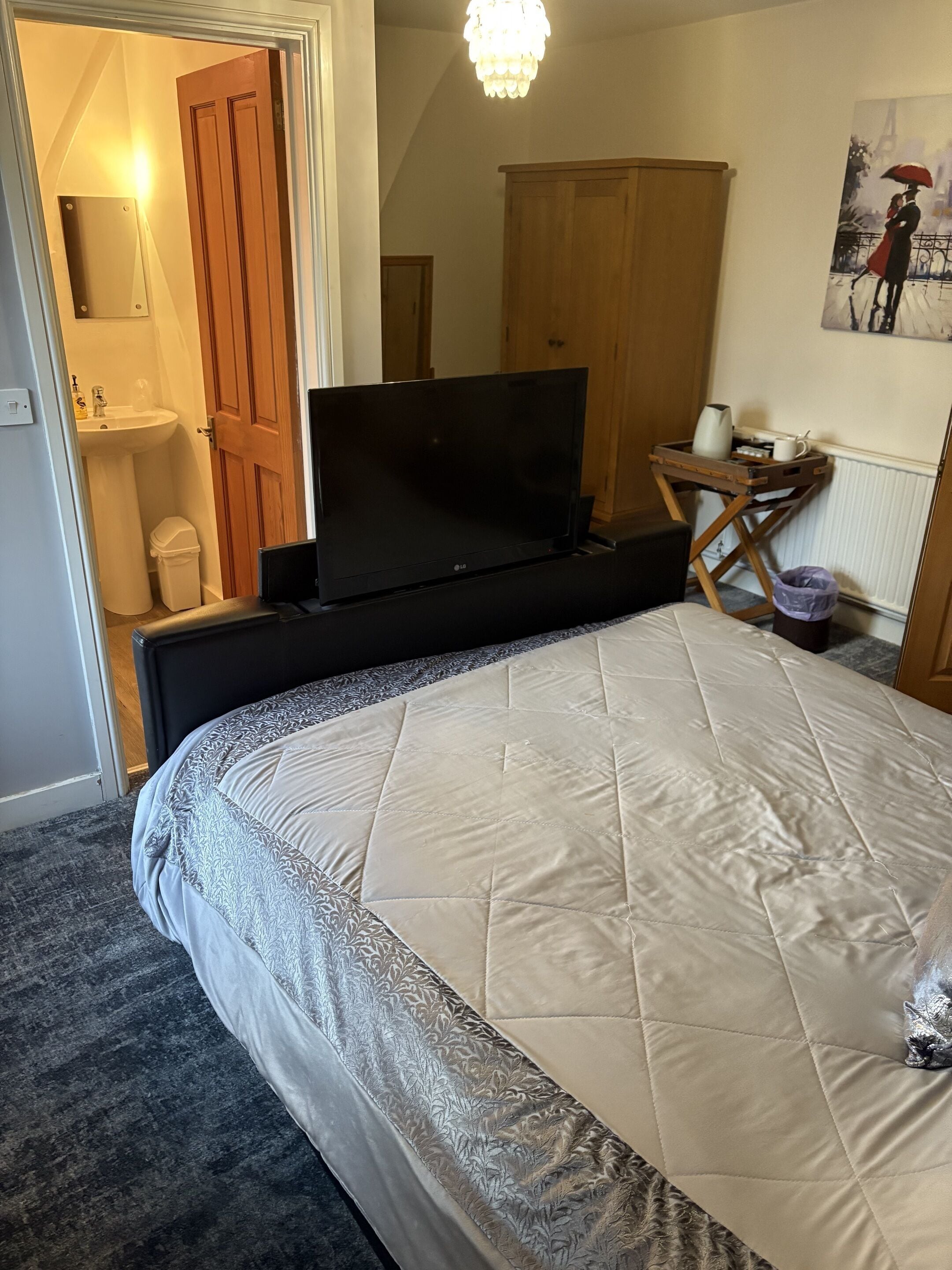 Quarto com cama, televisão, uma pequena mesa com chaleira e um quadro na parede.