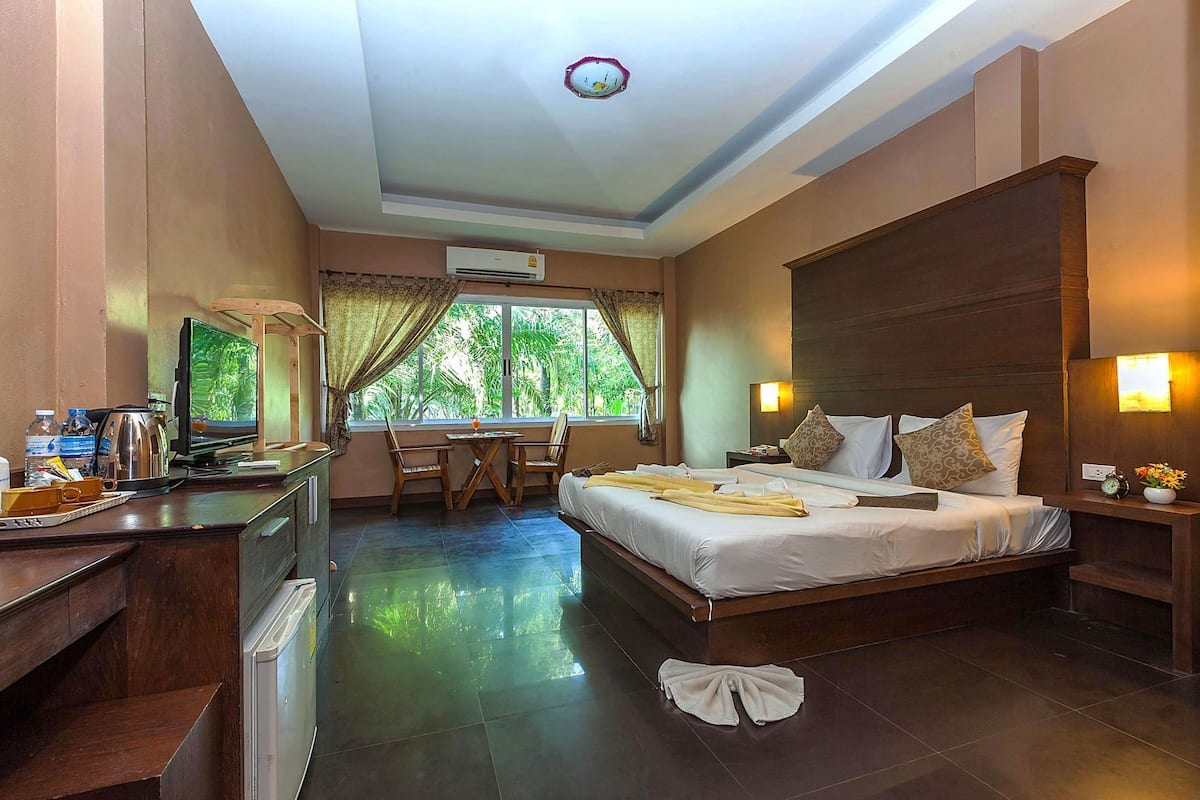 Quarto de hotel com cama, mesas de cabeceira, escrivaninha com televisão, janela com cortinas e vista para o verde lá fora.