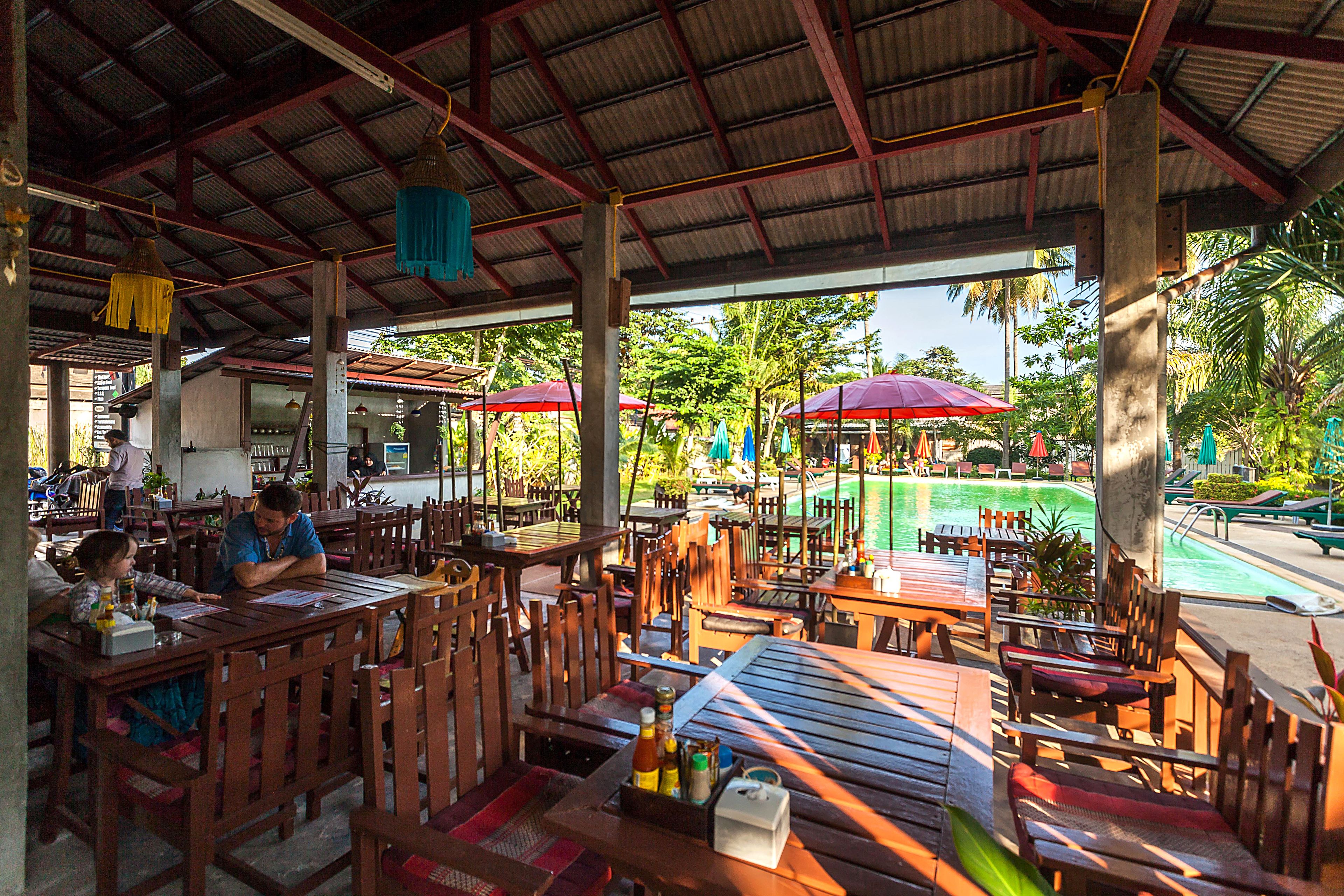 Restaurante ao ar livre com mesas e cadeiras de madeira, guarda-sóis e vista para uma piscina.