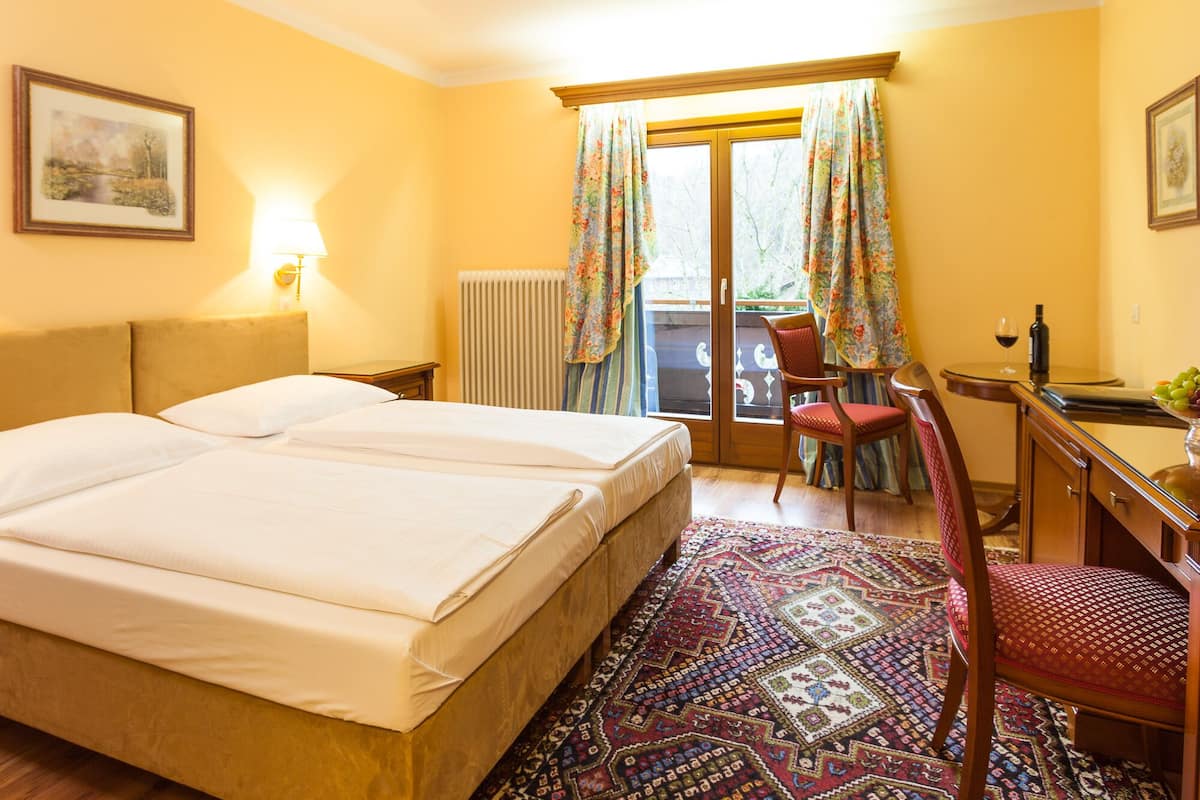 Quarto de hotel com duas camas, uma escrivaninha com cadeira, um radiador e uma janela com cortinas.