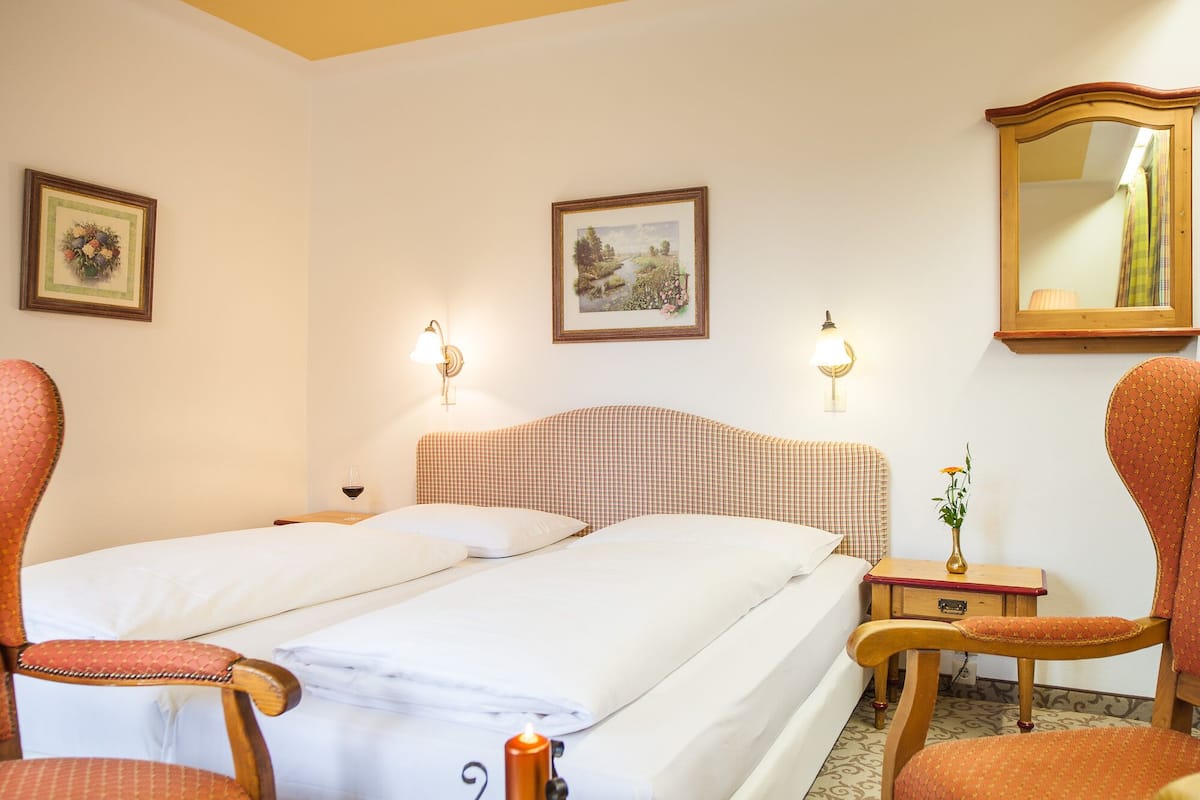 Quarto de hotel com duas camas, cabeceira de madeira, duas luminárias fixadas na parede, um espelho e uma pequena mesa com vela e frutas.