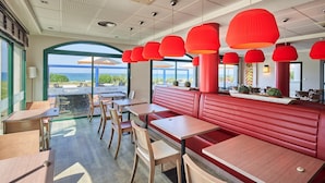 Um restaurante com assentos e mesas vermelhas, vista para o mar e luminárias pendentes vermelhas.