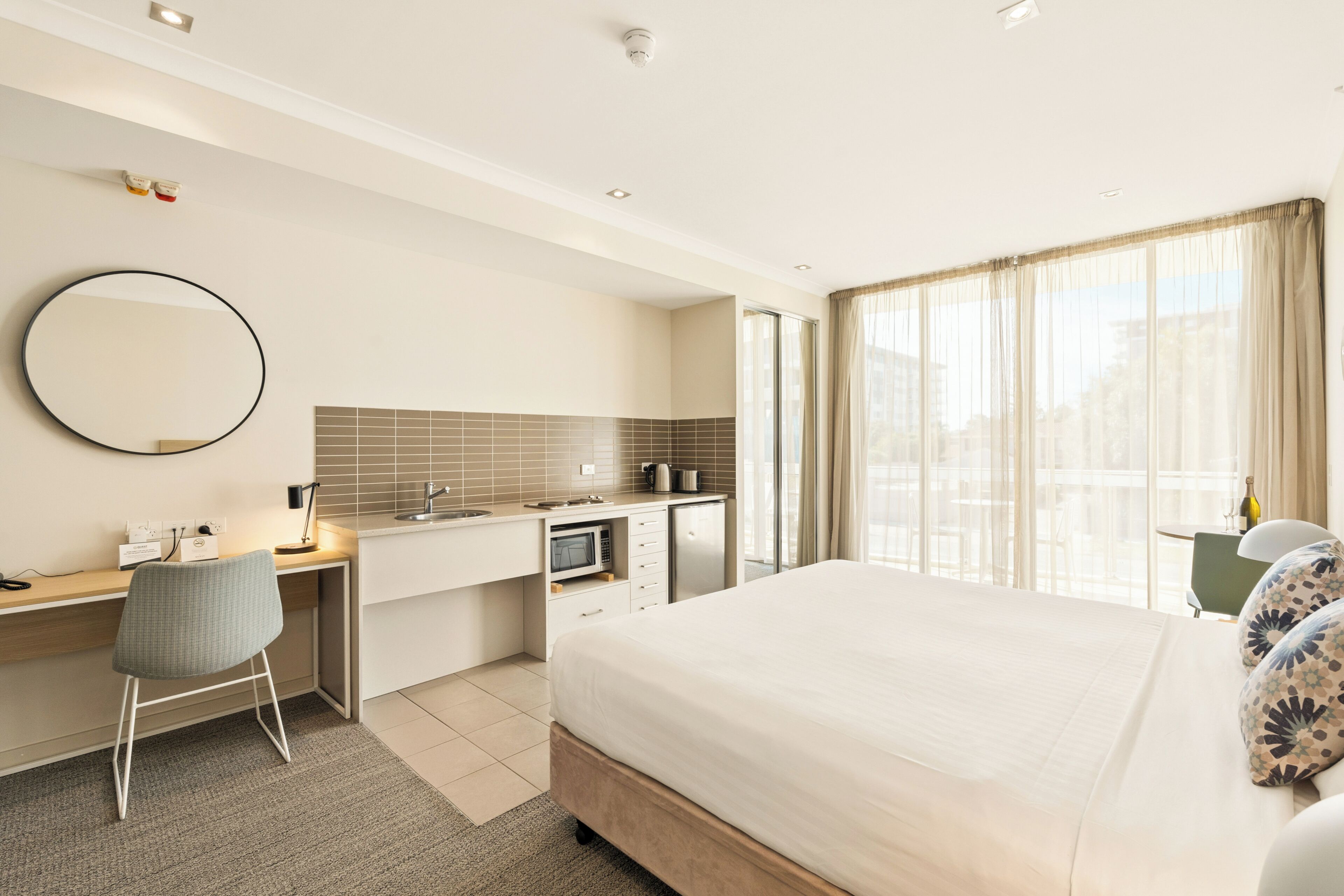 Quarto de hotel com cama, secretária, cadeira e cozinha compacta.