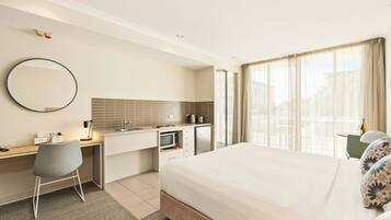 Quarto de hotel com cama, secretária, cadeira e cozinha compacta.