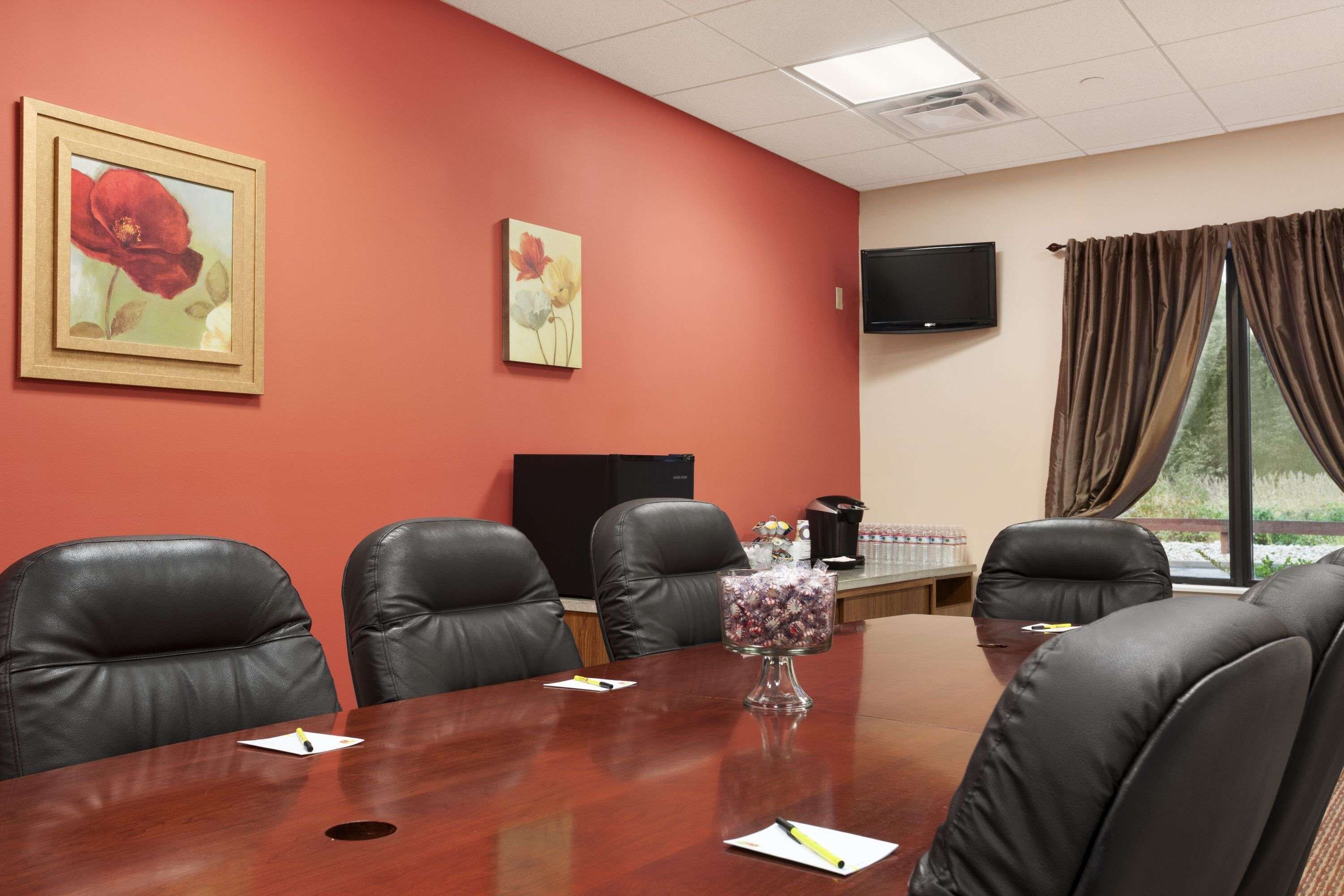 Uma sala de conferências com uma mesa de madeira, quatro cadeiras de couro preto, um vaso de vidro com flores e dois quadros florais na parede.
