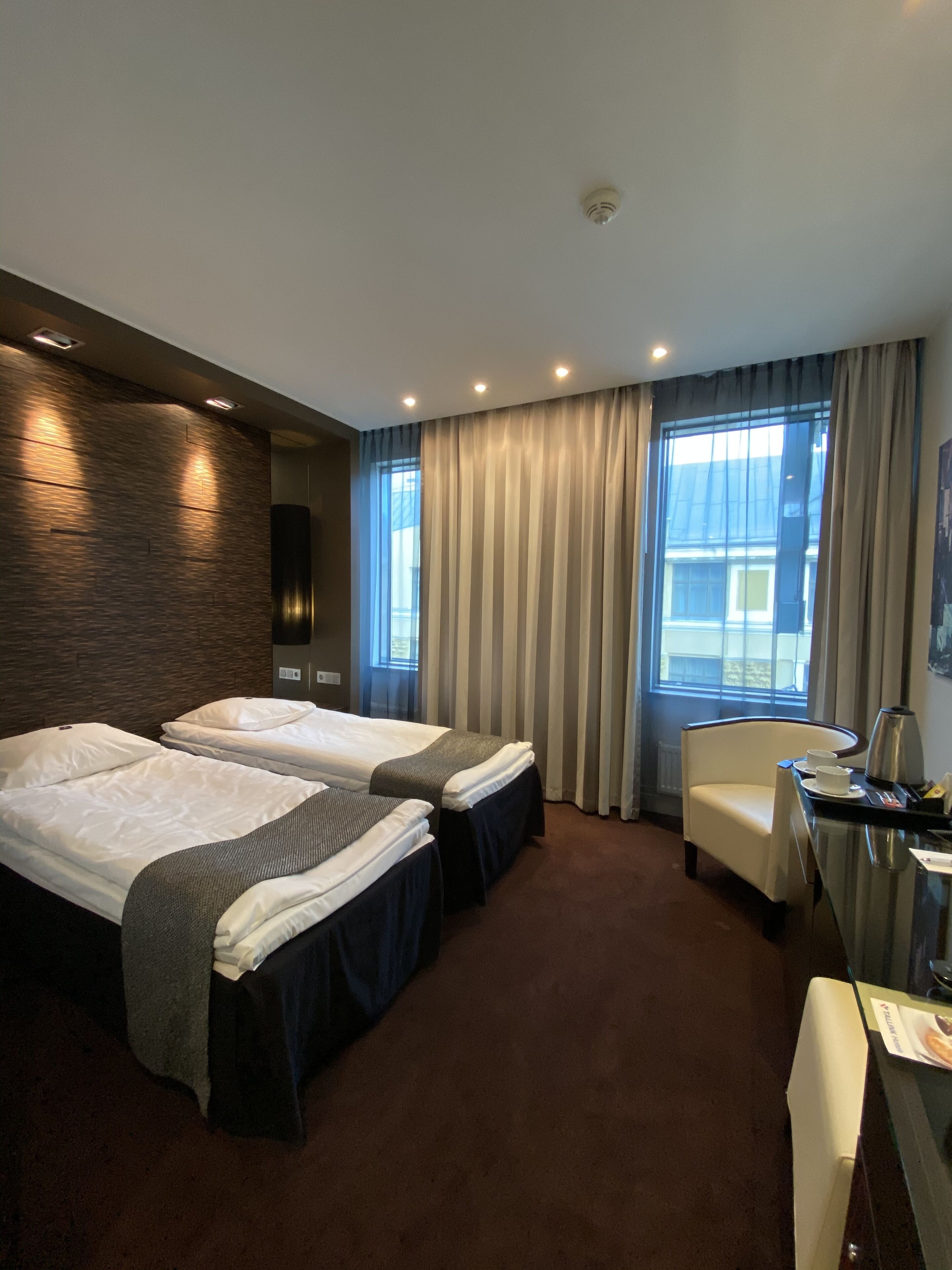 Quarto de hotel com duas camas, uma escrivaninha, uma cadeira e uma janela grande com cortinas.