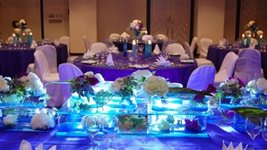 Salão de banquetes com mesas redondas preparadas para um evento formal, com copos de vidro com tonalidade azul e arranjos florais.