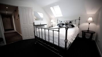 Um quarto com estrutura de cama em metal, uma mesa de cabeceira com abajur, uma janela grande e uma porta que dá para outro cômodo.