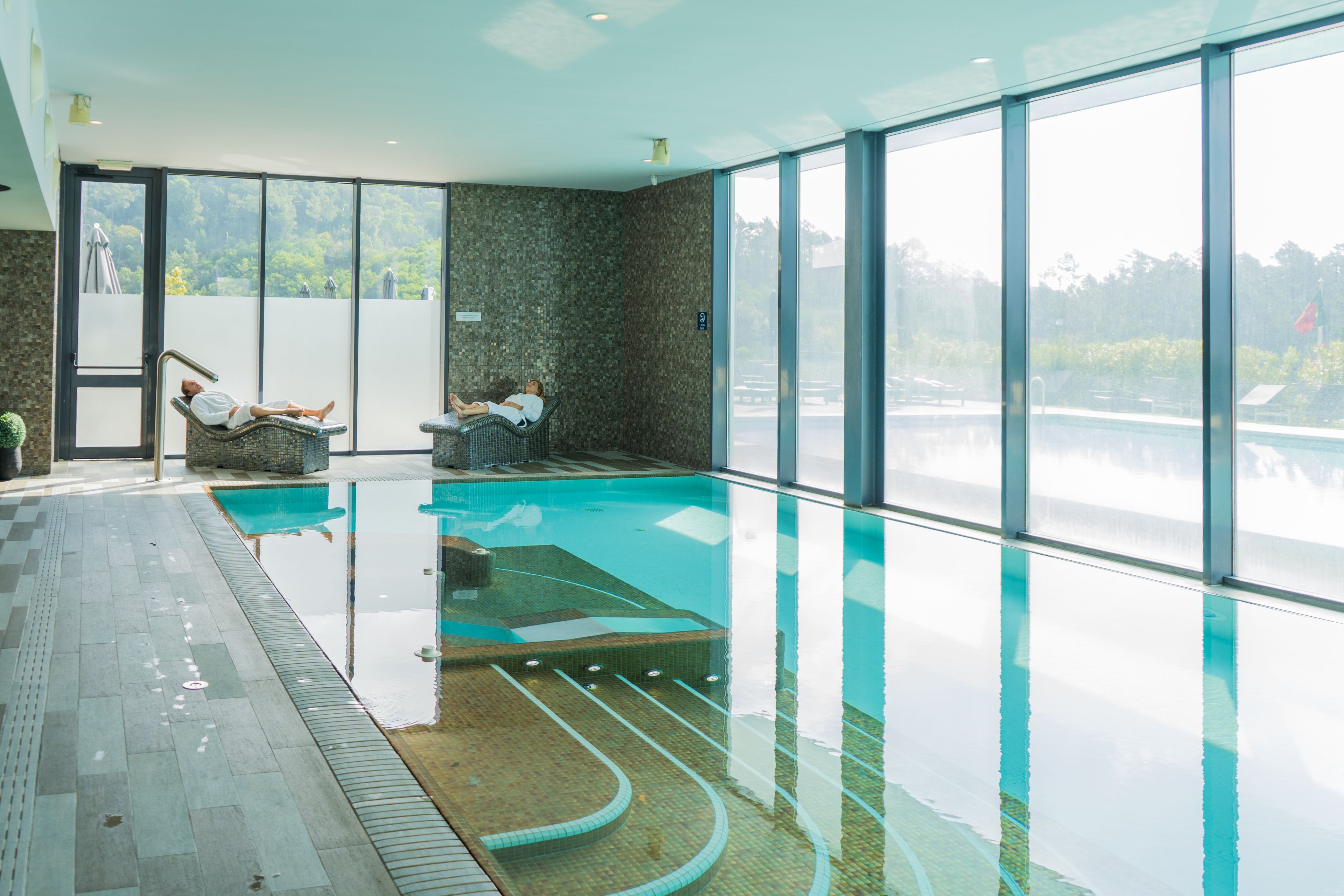Piscina interior com paredes de vidro transparente, chão revestido de azulejos e duas cadeiras de descanso.