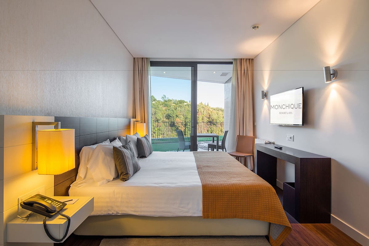 Quarto de hotel moderno com cama, abajures, escrivaninha com telefone, televisão e vista para o exterior.