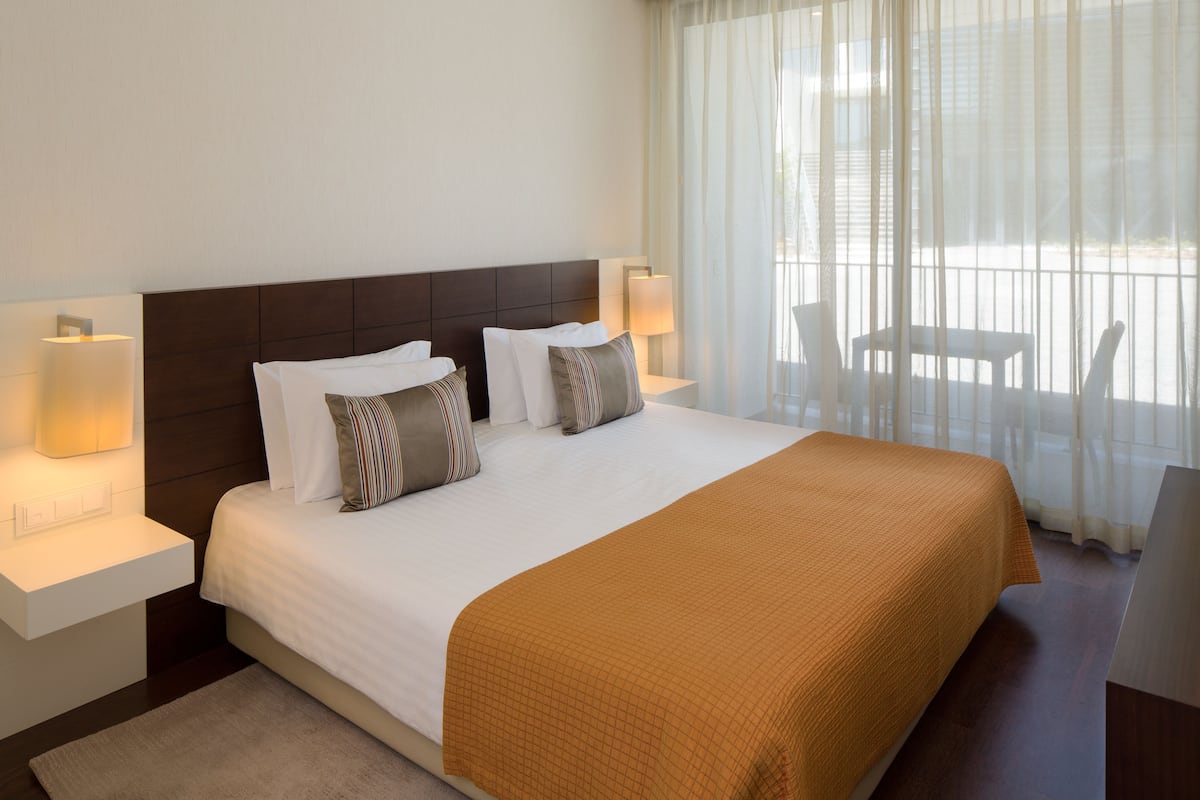 Quarto de hotel com uma cama grande, duas mesas de cabeceira com luminárias, vista para o exterior através de cortinas translúcidas, e uma pequena mesa e cadeira junto à janela.