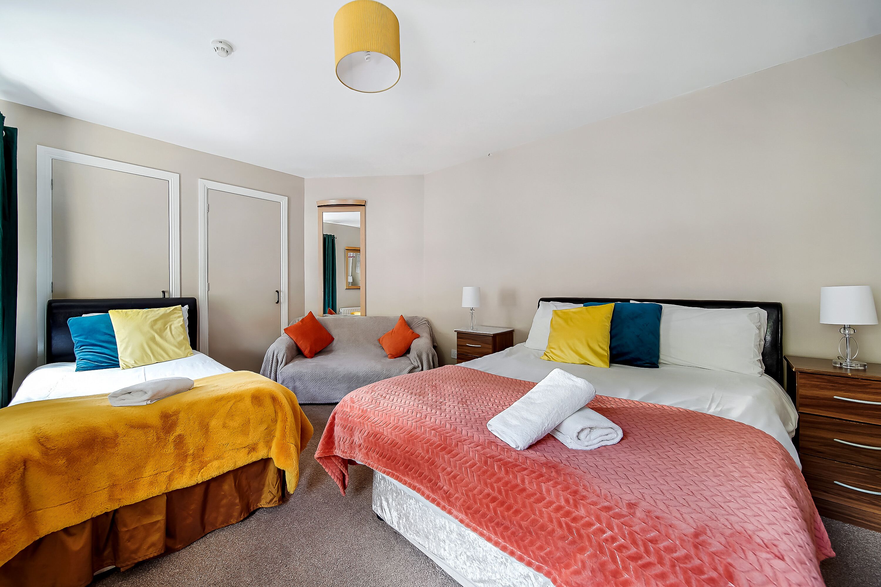 Quarto de hotel com duas camas, um sofá, um espelho e uma luminária.