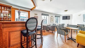 Uma área de bar com balcão e bancos de madeira, um espaço de estar com mesas e cadeiras, e uma televisão presa na parede.
