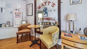 Um quarto com piso de madeira, uma poltrona de couro marrom, uma pequena mesa redonda, um abajur e um espelho com a inscrição 'WATT'S TYRCONNELL'.