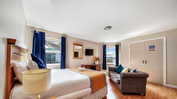 Quarto de hotel com cama, sofá, escrivaninha, televisão e janela com cortinas.