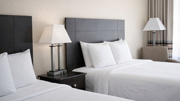 Quarto de hotel com duas camas, uma mesa de cabeceira com abajur e uma cadeira.
