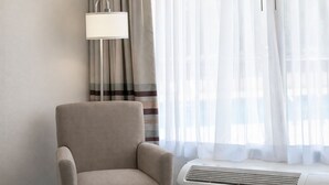 Quarto de hotel com uma poltrona, abajur, janela grande com cortinas leves e ar condicionado fixado na parede.
