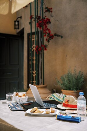 Uma mesa com um laptop, uma garrafa de água, um boné, um prato com pastelaria, um caderno e uma chávena.