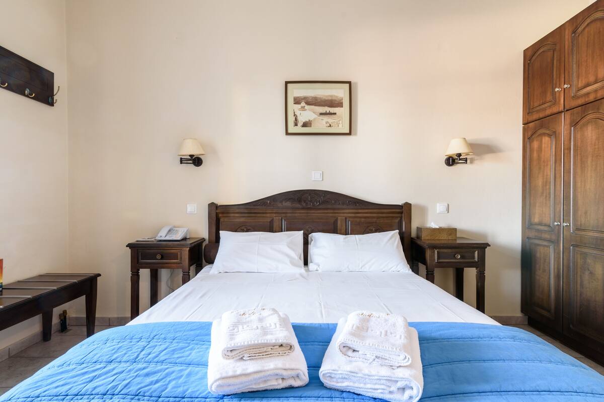 Cama bem arrumada com lençóis brancos e duas toalhas dobradas, cabeceira de madeira, mesas de cabeceira com luminárias e um quadro na parede.