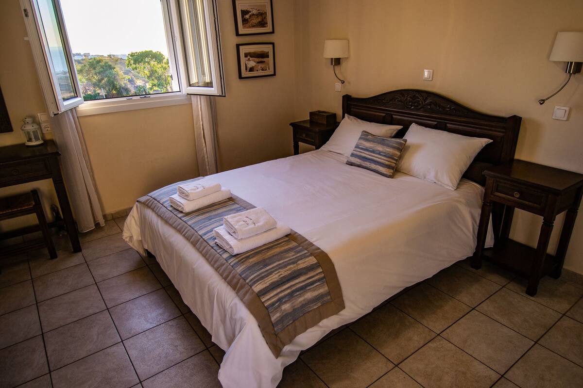 Uma cama bem arrumada com manta e travesseiros listrados, uma mesa de cabeceira com abajur, uma janela com cortinas e um quadro na parede.