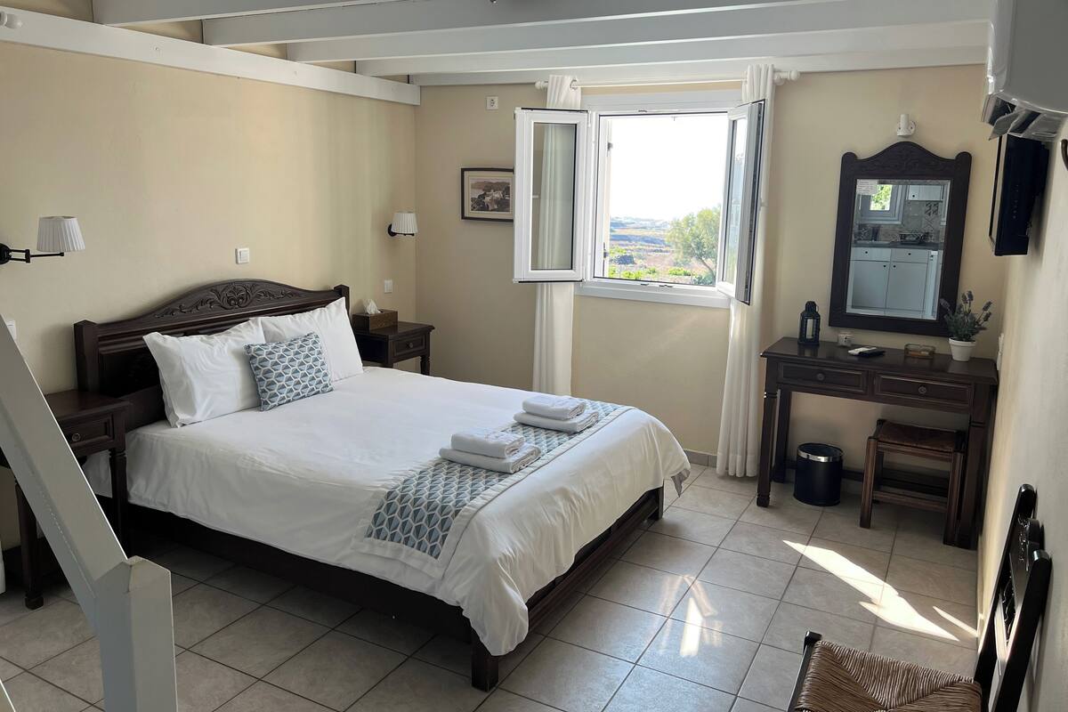 Cama bem arrumada com lençóis brancos, cabeceira de madeira e vista para o exterior através de uma janela aberta.