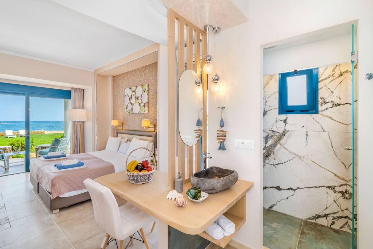 Quarto de hotel com cama, escrivaninha com fruteira, bacia de pedra e banheiro com parede de mármore e cortina de chuveiro azul.