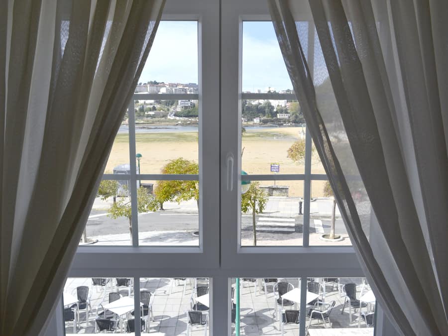 Visão pela janela com cortinas, mostrando uma área externa com mesas e cadeiras, um campo gramado e edifícios ao fundo.
