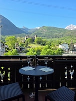 Varanda com duas mesas, cada uma com uma taça de vinho e um recipiente de metal, com vista para uma paisagem montanhosa que inclui um castelo.