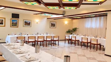 Uma sala de banquetes com mesas compridas preparadas para refeições, vigas de madeira no teto e quadros decorando as paredes.