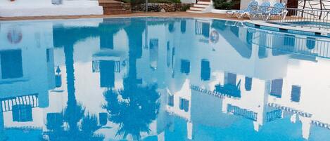 Uma piscina com água límpida refletindo a arquitetura ao redor. Há espreguiçadeiras e um salva-vidas à beira da piscina.
