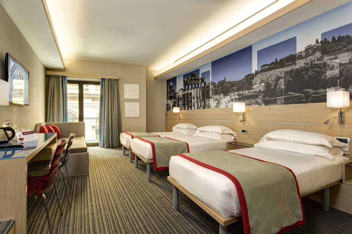 Quarto de hotel com três camas, uma escrivaninha com cadeira, televisão e um mural grande na parede.