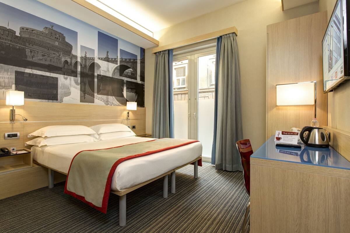 Quarto de hotel com uma cama, mesas de cabeceira, escrivaninha, chaleira e um grande mural na parede.