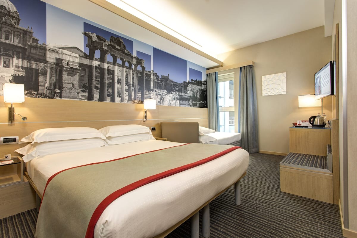 Quarto de hotel com uma cama grande, uma escrivaninha com luminária, televisão e vista para a cidade.