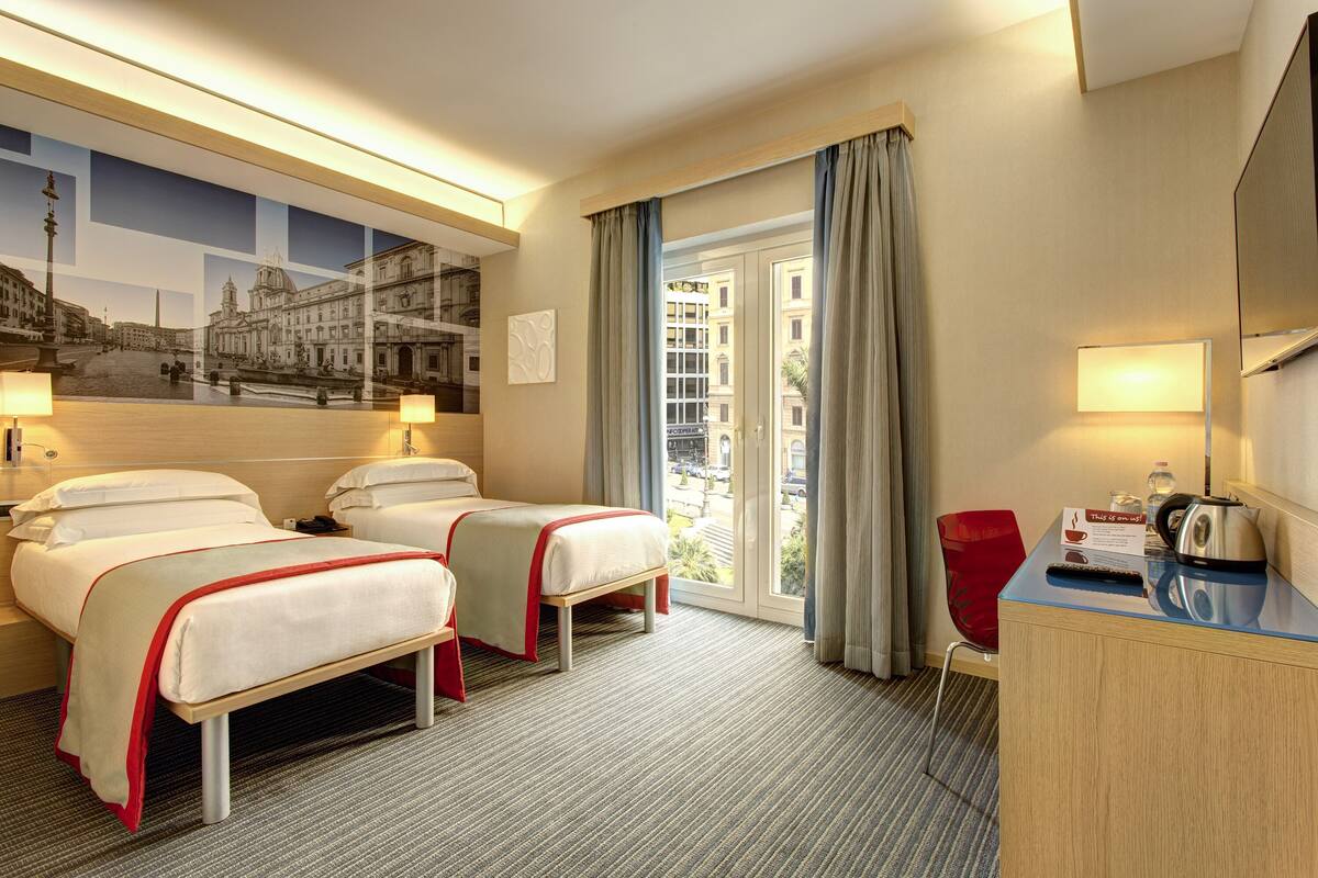 Quarto de hotel com duas camas, uma escrivaninha, uma cadeira, uma janela grande com cortinas e vista para a paisagem urbana.