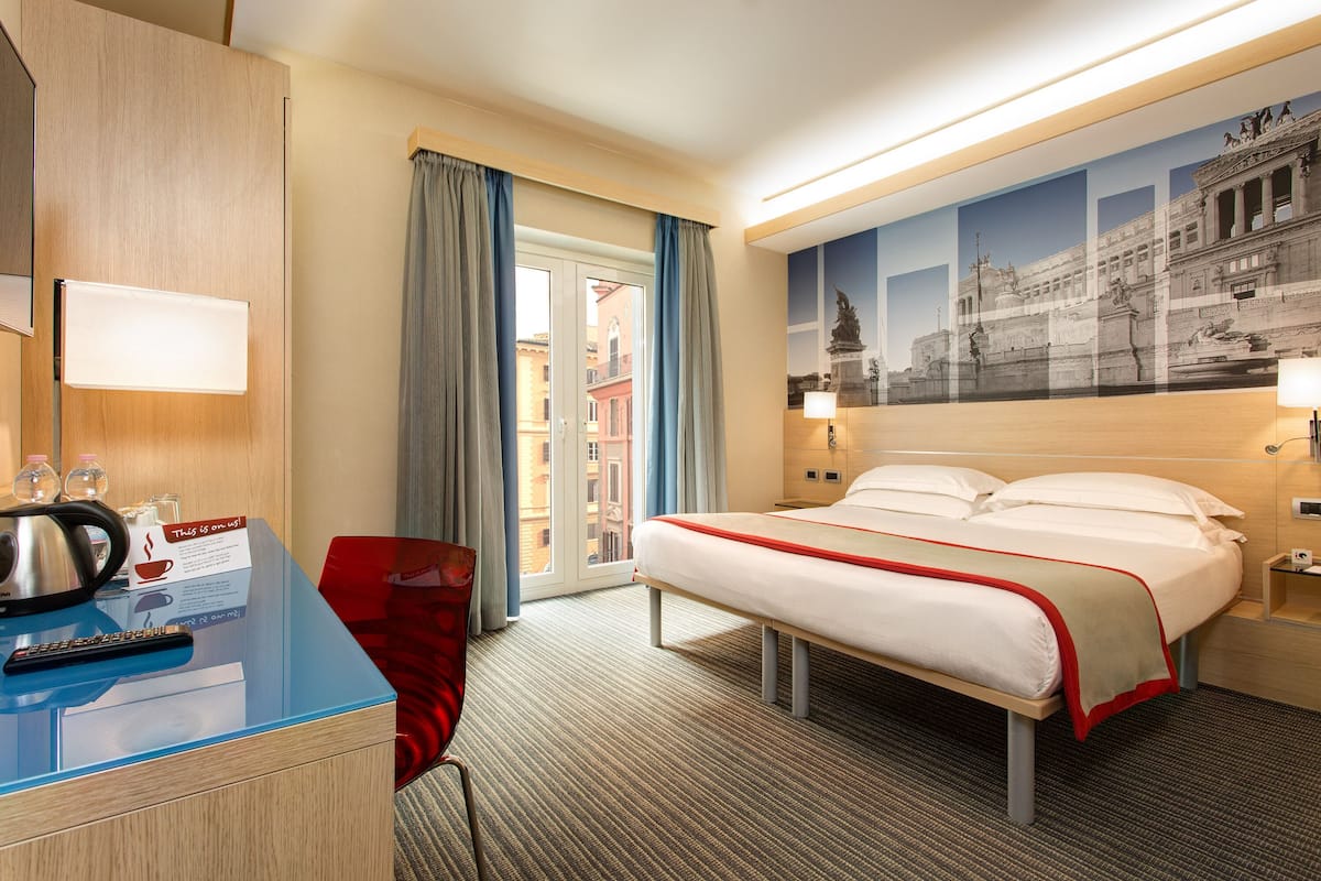 Quarto de hotel com cama, escrivaninha com cafeteira, cadeira, janela com cortinas e parede com mural da paisagem urbana.