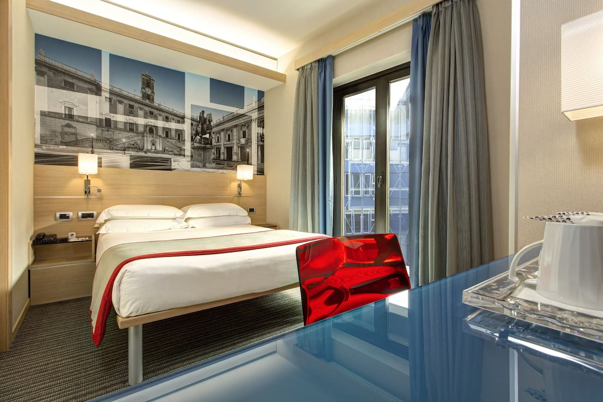 Quarto de hotel com cama, mesa de cabeceira, luminária, janela com cortinas e parede com mural da paisagem urbana.