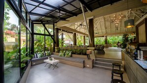 Um espaço interno amplo com parede de vidro, área de estar com almofadas, mesa e balcão de bar.