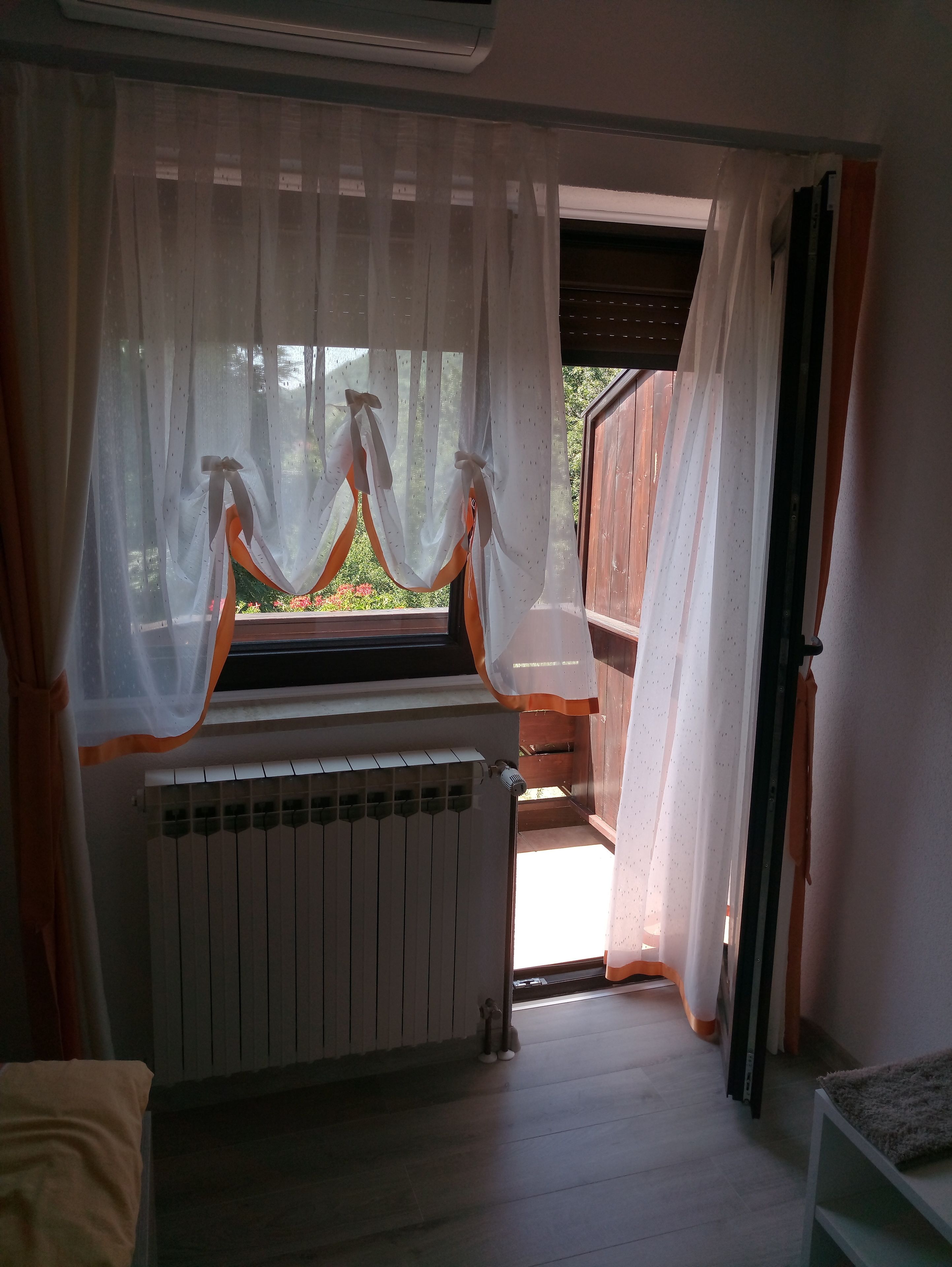 Quarto com radiador, janela com cortinas leves e fitas laranja, e vista para o verde lá fora.