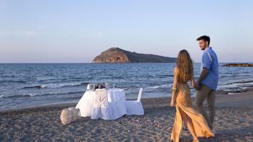 Cenário de praia com uma mesa posta para dois, uma cadeira e um objeto decorativo na areia.