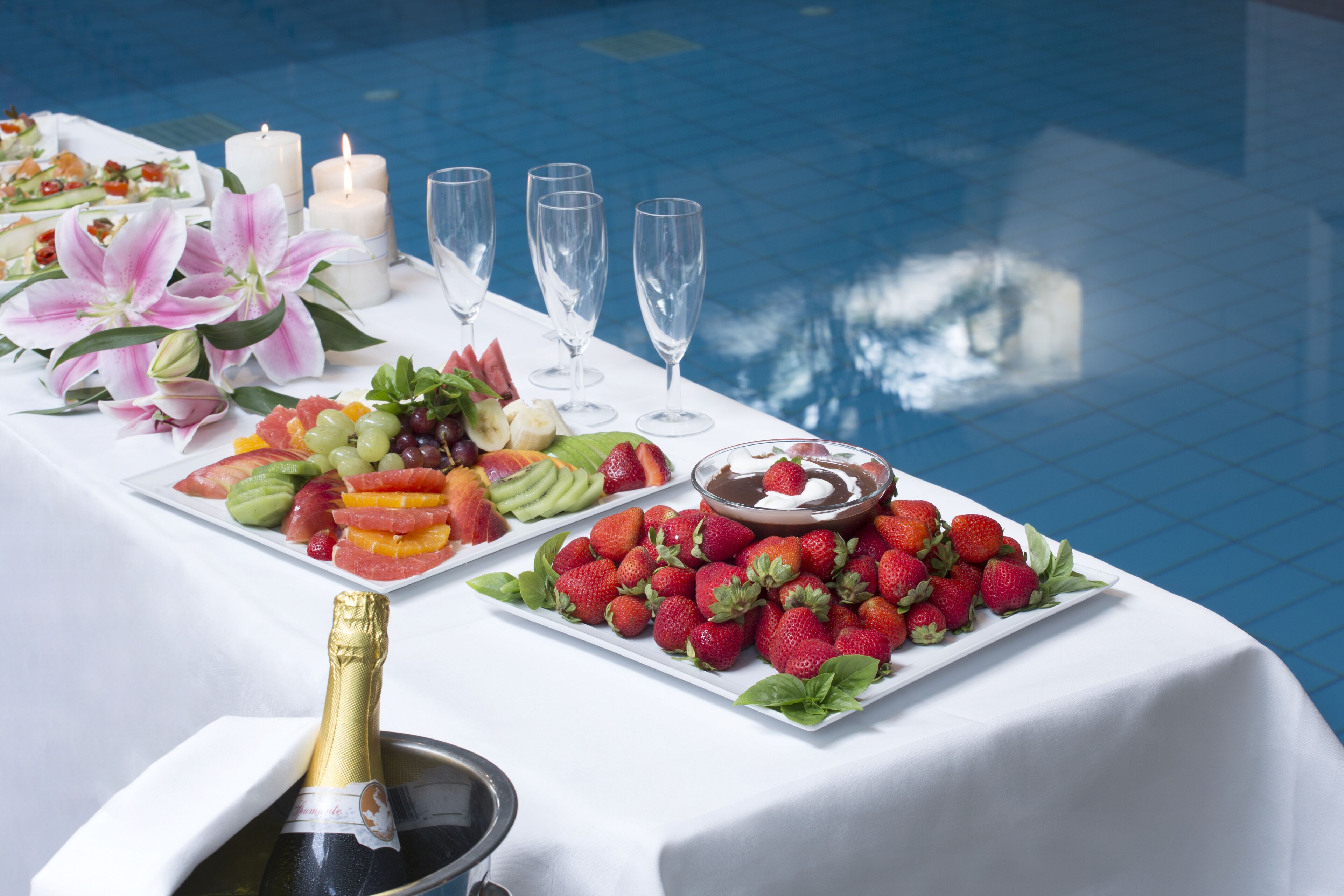 Uma mesa com diversas frutas e uma garrafa de champanhe, com uma piscina ao fundo.