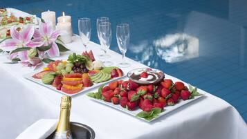 Uma mesa com diversas frutas e uma garrafa de champanhe, com uma piscina ao fundo.