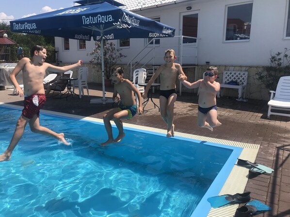 Uma piscina com quatro pessoas pulando. Há cadeiras de descanso e um guarda-sol azul com a marca 'Natura Aqua' visível.