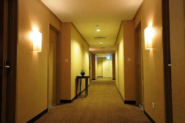 Corredor de hotel com piso em carpete, luminárias fixadas na parede e uma porta no final.