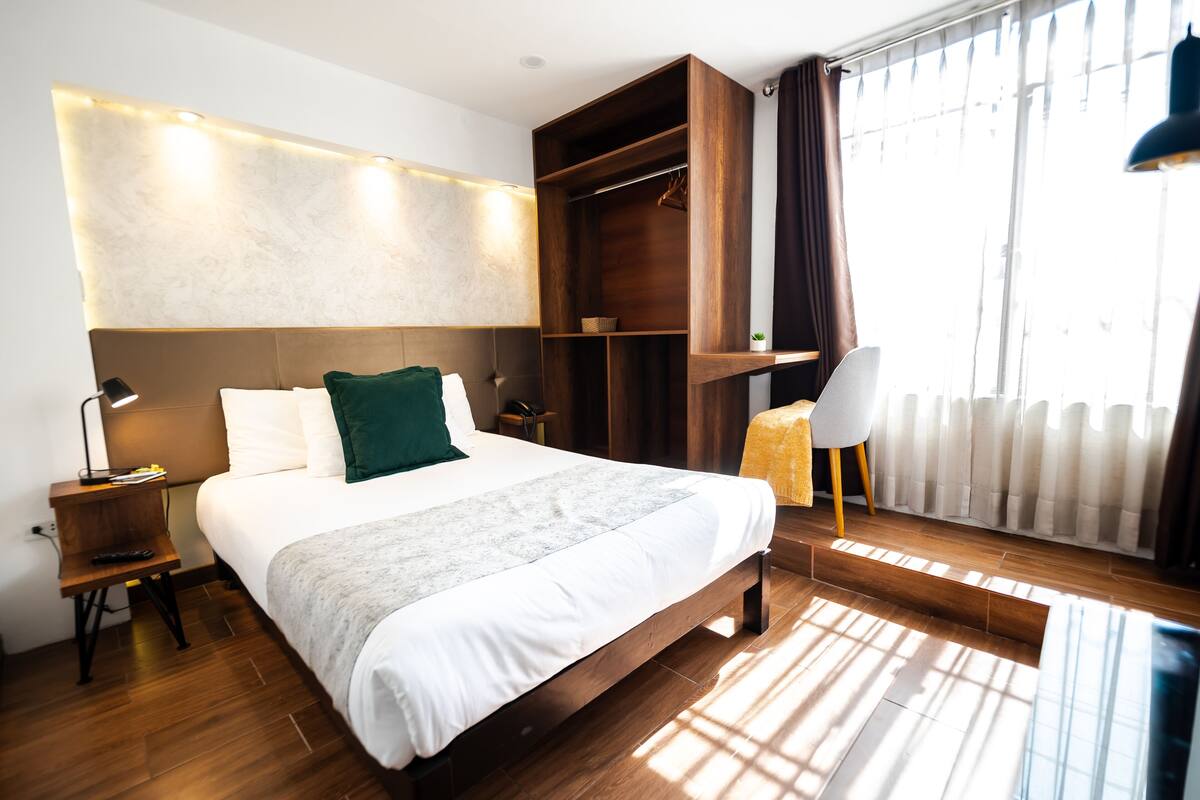 Quarto de hotel moderno com uma cama grande, mesa de cabeceira, guarda-roupa e janela com cortinas.