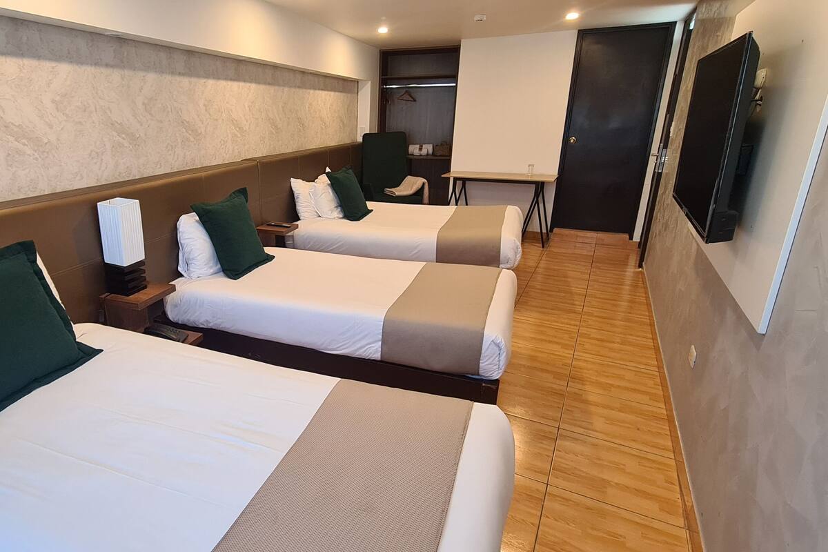 Quarto de hotel com três camas, uma televisão, uma escrivaninha e uma cadeira.