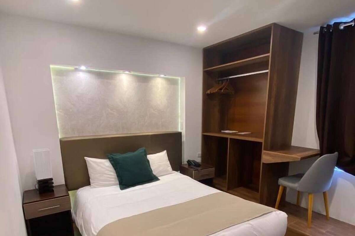 Quarto de hotel com cama, mesa de cabeceira, escrivaninha, cadeira e armário embutido.