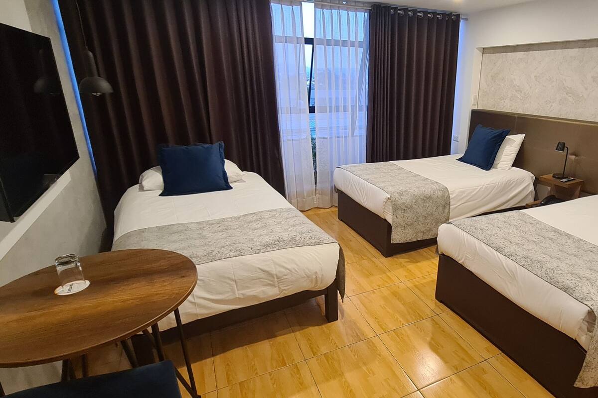 Quarto de hotel com duas camas, uma mesinha e vista pela janela com cortinas.