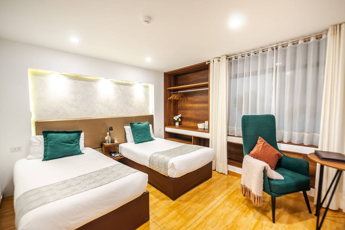Quarto de hotel com duas camas, cabeceira de madeira, uma poltrona verde, uma mesinha e uma janela com cortinas.