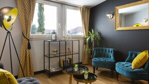 Uma sala de estar com sofá, uma poltrona azul, uma mesinha com plantas, estante de livros e uma janela grande com cortinas.